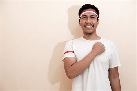 독립기념일을 축하하며 흰 티셔츠를 입고 머리와 팔에 빨간색과 흰색이 섞인 넥타이를 매고 한 손은 가슴에 얹고 미소 짓고 있는 동남아시아 남성 17에 대한 스톡 사진 및 기타