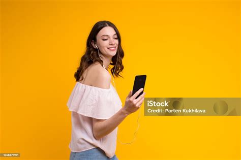 세련된 여름 블라우스 옷에 행복 귀여운 곱슬 갈색 머리 소녀 노란색에 고립 된 서 스마트 폰에 음악 응용 프로그램 다운로드에 듣고 오디오 노래를 선택하는 즐기는 현대 이어폰