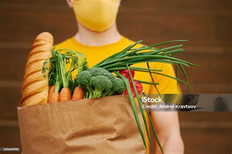 노란색 유니폼 모자 티셔츠 얼굴 마스크 장갑을 입은 식료품 배달 남자는 나무 배경에 음식 과일 채소가있는 종이 가방을 들고