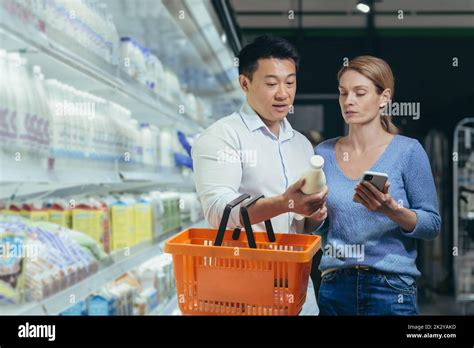Young Happy Asian Couple Using Smartphone In Supermarket With Shopping Cart Choosing Products