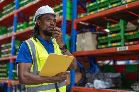 조끼와 헬멧을 착용한 흑인 남성 창고 노동자가 휴대용 무전기를 이용해 창고 공장 매장에서 물건을 확인하면서 동료와 통화하고 있다 물류 물류센터 개념 30 34세에 대한 스톡
