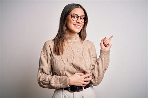 젊은 아름 다운 갈색 머리 사업가 캐주얼 스웨터와 안경 얼굴에 큰 미소와 함께 서 서 카메라를 보고 측면에 손과 손가락으로 가리키는 가리키기에 대한 스톡 사진 및 기타