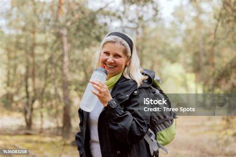 숲 속의 중년 여성이 친구들과 즐겁게 지내며 웃고 있습니다 손에 물병을 들고 마시는 스마트 시계를 가진 현대 노인 엄마 히킹 캠핑 트레킹 미소에 대한 스톡 사진 및 기타