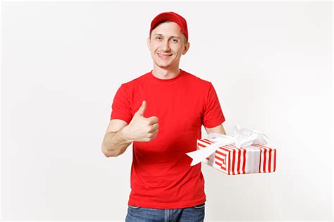 흰색 배경에 고립 된 빨간 유니폼에 배달 남자 남성 모자에 웃 고 T셔츠 청바지로 택배 또는 대리점 빨간 줄무늬 선물 상자를 들고 일하고 제시 광고에 대 한 공간을 복사