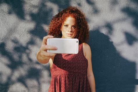 얼굴에 주 근 깨와 매력적인 빨간 머리 여자는 화창한 날 야외에서 회색 배경에 서 있는 동안 스마트폰 카메라에 의해 Selfie가 하고있다 사진의 허리 곱슬 머리에 대한