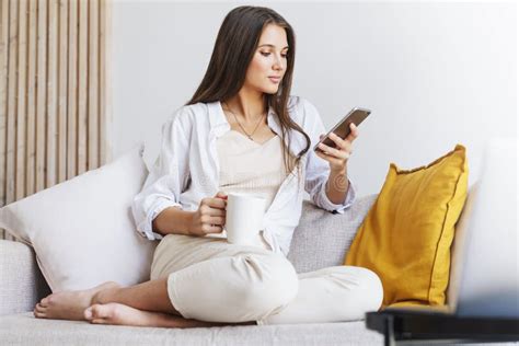 Улыбаясь красивая блондинка в белой рубашке сидит на диване с белой Easydate смартфонов на чашке