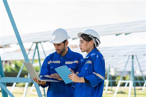 태양 전지판 전원 농장에서 일하는 노트북 컴퓨터를 들고 있는 두 명의 남성과 여성 전기 기술자가 태양광 태양 전지 패널의 긴 행 배열 사이에 있습니다 녹색 에너지와 청정