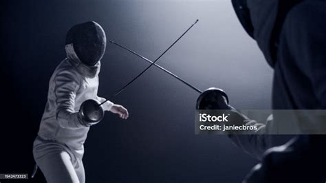 두 명의 전문 펜싱 선수가 전투에서 서로 마주보고 있는 검을 휘두르며 장엄한 파란색 무대 조명과 연기 효과를 연출합니다 낮은 각도보기 칼 교차 펜싱에 대한 스톡 사진 및