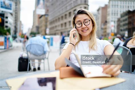 시력 보호를 위한 광학 안경을 쓴 중국 여성은 거리 카페에서 여가 동안 계획 조직을 요구하고 밀레니엄 소녀는 스마트폰을 통해 통신하면서 교과서에 메모를 하고 있다 20 24