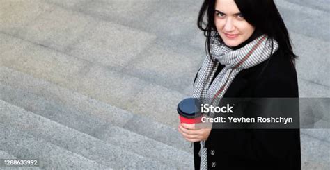 코트와 스카프를 입은 젊은 세련된 여성이 가을 공원의 야외에서 빨간색 에코 종이 유리에서 아침 뜨거운 커피를 마십니다 테이크