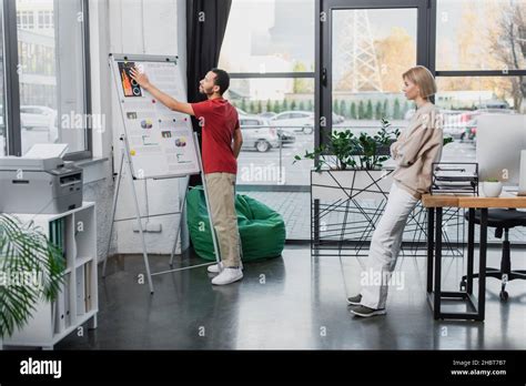 Full Length Of African American Man Pointing At Charts On Flip Chart Near Blonde Colleague Stock