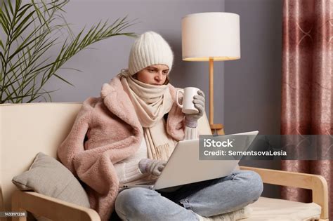 모자 장갑 스카프 코트를 입고 뜨거운 차 한 잔과 함께 집 안에 앉아있는 화가 난 얼어 붙은 여성의 사진 워밍업을 시도하고