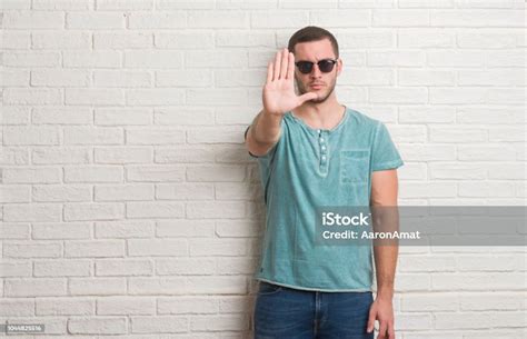 젊은 백인 남자 오픈 손으로 심각 하 고 자신감 표현 방어 제스처 정지 표시를 하 고 선글라스를 착용 하는 흰색 벽돌 벽을 통해 서 경계 표지에 대한 스톡 사진 및 기타