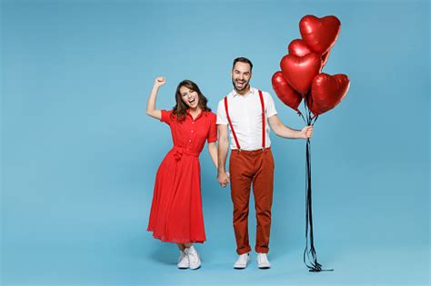 생일 휴가 파티를 축하하는 흰색 빨간 옷을 입은 행복한 젊은 부부 친구 남자 여성의 전체 길이는 파란색 배경에 고립 된 우승자 제스처를하는 공기 팽창 헬륨 풍선의 무리를