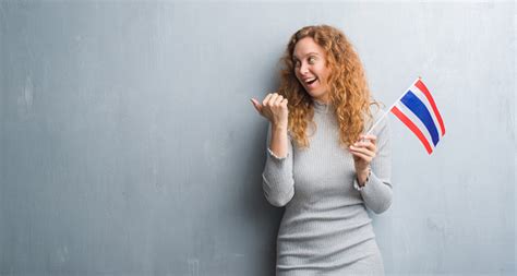 태국의 국기를 들고 회색 그런 지 벽에 젊은 빨강 머리 여자는 고 행복 얼굴 웃 고 옆까지 엄지 표시 가리키기에 대한 스톡 사진 및 기타 이미지 Istock