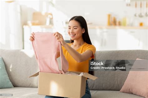 소파에 앉아서 판지 상자를 열고 배달을 확인하고 새 옷을 들고 웃는 것에 만족한 아시아 여성 개념에 대한 스톡 사진 및 기타