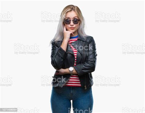 젊은 금발 여 자가 입고 패션 자 켓과 선글라스와 격리 된 배경 위에 질문 잠겨있는 표현에 대 한 생각 하는 턱에 손을 사려깊은