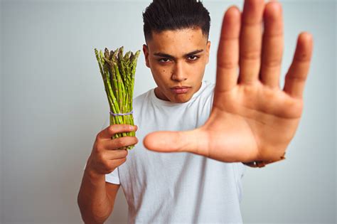 고립 된 회색 배경 위에 서있는 아스파라거스를 먹는 젊은 브라질 남자는 진지하고 자신감있는 표정 방어 제스처로 정지 표지판을하는 열린 손으로 서 있습니다 건강한 생활방식에