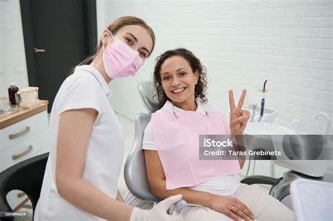 치과 의사 옆에 치과 의사 의자에 앉아 평화 표지판을 보여주는 행복한 아프리카 계 미국인 여성 환자 치과 치료를 받은 후 카메라를 보고 아름다운 이빨 미소로 미소 2명에