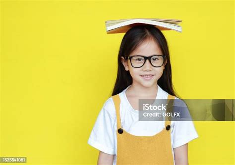 웃고 있는 아시아의 어린 소녀는 머리에 책이 있는 안경을 쓰고 복사 공간이 있는 노란색 배경에 고립된 카메라를 보고 있다 학생과