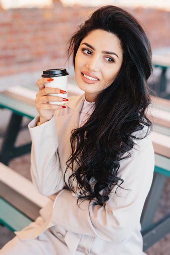어두운 머리 눈썹과 잘 생긴 입술을 옆으로 보고 야외 벤치에 앉아 테이크 아웃 컵에 카푸치노를 마시는 미소와 화려한 여자