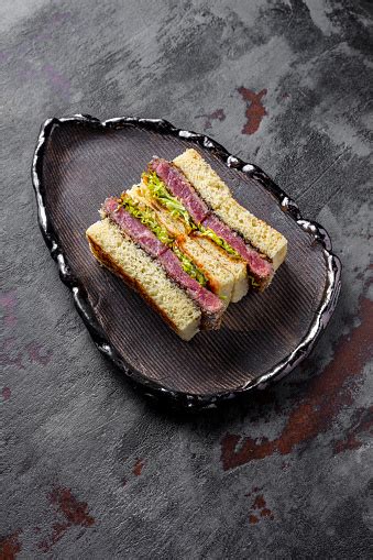 와규 일본식 쇠고기 스테이크 양상추 망고 칠리 소스를 곁들인 콘브레드 샌드위치 샌드위치는 나무 잎 형태의 세라믹 접시에 놓여 있습니다 접시는 청동색이 튀는 회색 돌 배경에