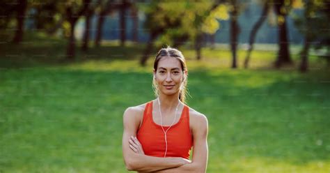 운동복을 입고 포니 테일로 팔짱을 끼고 초원에 서있는 건강한 슬림 미소 백인 갈색 머리에 적합합니다 귀에는 이어폰이 있습니다