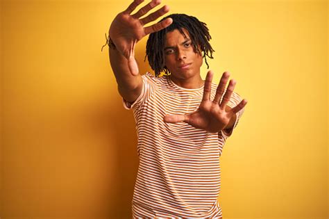 손바닥과 손가락을 사용하여 프레임을하고 고립 된 노란색 배경 위에 서있는 줄무늬 티셔츠를 입은 공포를 가진 아프로 남자 카메라