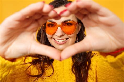 스웨터 블루 바지 스튜디오에서 노란색 주황색 배경에 고립 된 손으로 모양 마음을 보여주는 선글라스의 매력적인 젊은 여성 사람들은 진심으로 감정 라이프 스타일 개념입니다 복사