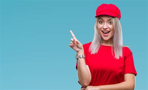 젊은 금발 여자 얼굴에 큰 미소와 함께 고립 된 배경 위에 빨간 모자를 쓰고 카메라를 보고 옆에 손 및 손가락으로 가리키는 가리키기에 대한 스톡 사진 및 기타 이미지