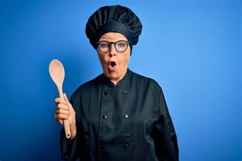 수석 아름 다운 회색 머리 요리사 여자 밥 솥 유니폼과 모자를 들고 나무 숟가락을 들고 깜짝 얼굴 두려움과 흥분 노인에 대한