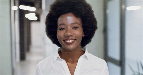 현대적인 사무실 건물 안에 서서 흰색 셔츠를 입고 카메라를 바라보며 아프리카 머리를 한 예쁜 웃는 흑인 젊은 여성의 얼굴 초상화를 닫습니다 사업가 실내 초상화 사진