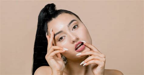 포니테일 헤어스타일을 가진 아름 다운 젊은 섹시한 아시아 여성 베이지 색 배경에 고립 된 얼굴 입술과 눈썹을 만지는 손으로 카메라에 포즈를 취합니다 인물 사진을