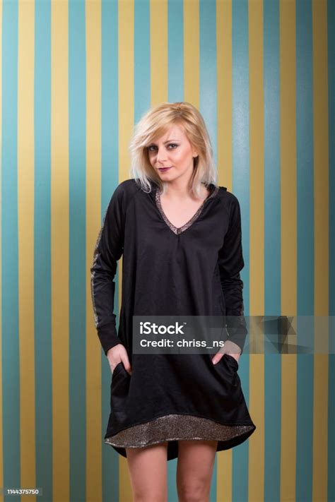매력적인 젊은 금발의 여자 앞에서 포즈 줄무늬 노란색 파란색 배경 착용 다크 드레스 손에 주머니에 미소와 카메라를 보고 가냘픈에 대한 스톡 사진 및 기타 이미지 Istock