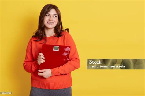 오렌지 캐주얼 스웨터를 입은 잘 생긴 갈색 머리 여성은 바닥 비늘을 포용하고 꿈꾸는 표정으로 멀리 바라보고 노란 배경을 통해 고립 된 홍보 공간을 복사합니다 20 24세에