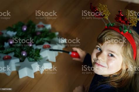 크리스마스 이브나 첫 강림에 강림절 화환에 촛불을 켜고 있는 재미있는 크리스마스 머리띠를 가진 어린 미취학 소녀 독일의 전통 화환 가족 휴가를 축하하는 행복한 아이 강림절에