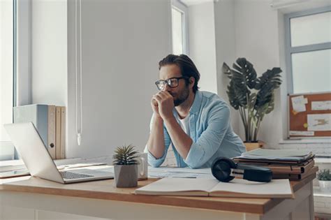 걱정스러운 청년이 사무실에 있는 자신의 직장에 앉아있는 동안 손에 머리를 숙이고 걱정하는에 대한 스톡 사진 및 기타 이미지
