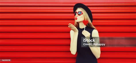 막대 사탕을 가진 아름다운 젊은 여성의 초상화는 빨간 하트 모양의 선글라스 배경에 검은 둥근 모자를 쓰고 달콤한 공기 키스를 보내는 립스틱으로 입술을 불고 있다 25 29
