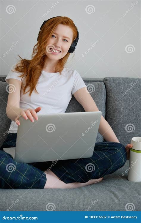 Redhead Girl In Pajamas And Headphones Sits On A Sofa With A Laptop On