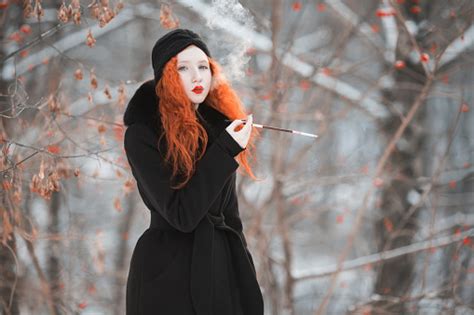 손에서 떠 있는 겨울 숲의 배경에 검은 코트에 붉은 머리를 가진 여자 담배와 그녀의 머리에 터 번으로 밝은 모습으로 빨간 머리
