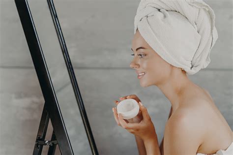 상쾌한 행복한 여성의 옆으로 촬영맨 어깨포즈 머리에 목욕 수건을 입고 거울로 자신을 보고 얼굴 크림을 적용 건강한 피부를 가지고 천연 화장품을 사용합니다 셀프 케어 컨셉