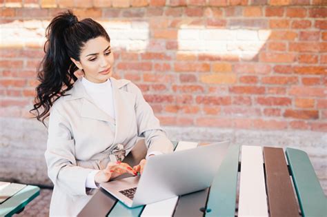인간과 기술 어두운 머리를 가진 젊은 예쁜 여자 조랑말 꼬리에 흰색 코트 에 옷을 입고 노트북에 입력 벽돌 벽을 통해 아늑한 카페에서 테이블에 앉아 소셜 네트워크를 통해