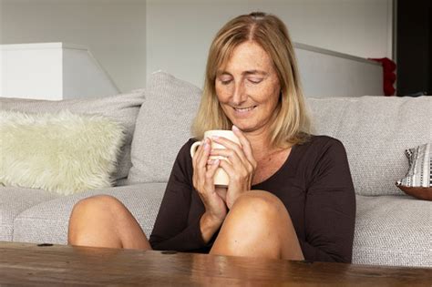 50대의 매력적인 주부 거실에서 잠시 멈춰 서서 차 한 잔과 함께 휴식을 취하며 과거와 미래의 삶에 대해 생각합니다 그녀는 거실에 혼자있다 가구에 대한 스톡 사진 및 기타
