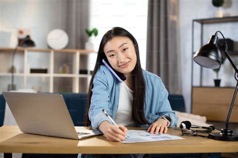 밝은 사무실이나 홈 인테리어에서 모바일로 이야기하면서 메모를 쓰는 웃는 아시아 여성 현대적인 노트북과 스마트폰을 사용하여 업무를 위해 검은 머리를 가진 예쁜 여성 사진