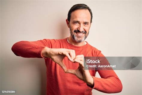 솔로 의외의 흰색 배경 위에 서있는 캐주얼 한 오렌지 스웨터를 입은 중년 남성은 손으로 심장 기호 모양을하고 사랑에 미소 로맨틱 한 개념 하트 모양에 대한 스톡 사진 및