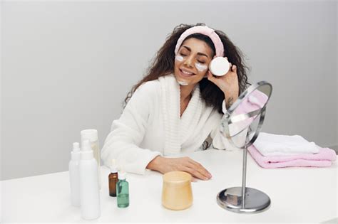 졸린 얼굴 크림을 들고 무두질한 예쁜 곱슬 라틴 아가씨가 미용 절차를 밟고 흰색 가정 인테리어의 의자에 앉아있는 것을 즐겼습니다 공간을 복사합니다 미용 의학 미용 제품 스킨