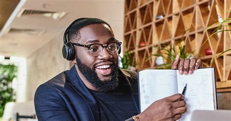 짙은 파란색 재킷과 안경을 쓴 긍정적인 세련된 젊은 아프리카계 미국인 사업가가 테이블에 앉아 실내 무선 이어폰을 착용하고 일반 노트북 컴퓨터에서 화상 회의를 통해 상사와