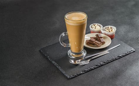 왼쪽 전망은 두 개의 머핀과 초콜릿 칩이 있는 아로마 커피 라떼를 숟가락과 포크가 있는 흰색 접시에 얹어 검은 돌 판에 얹어 놓습니다 0명에 대한 스톡 사진 및 기타 이미지