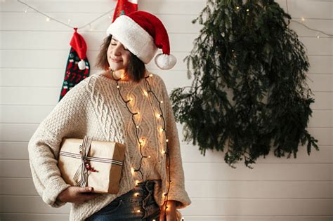 현대 크리스마스 트리 조명과 스타킹의 배경에 크리스마스 선물 상자를 들고 산타 모자에 세련된 행복 소녀 젊은 힙스터 여자 에 아늑한 스웨터 들고 선물 과 미소 12월에 대한
