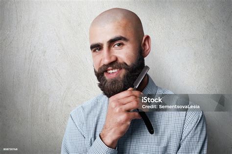 두꺼운 검은 수염과 콧수염 그의 수염에 면도칼을 들고 미소와 매력적인 캔디 Balded이 발사 젊은 미용사 입고 체크 셔츠 그의 면도칼의 날카로운 칼 날을 보여주는 날에